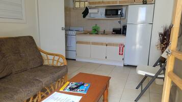 Um espaço compacto com sofá, uma mesa de madeira com um controle remoto, e uma área de cozinha compacta com micro-ondas, fogão e geladeira.