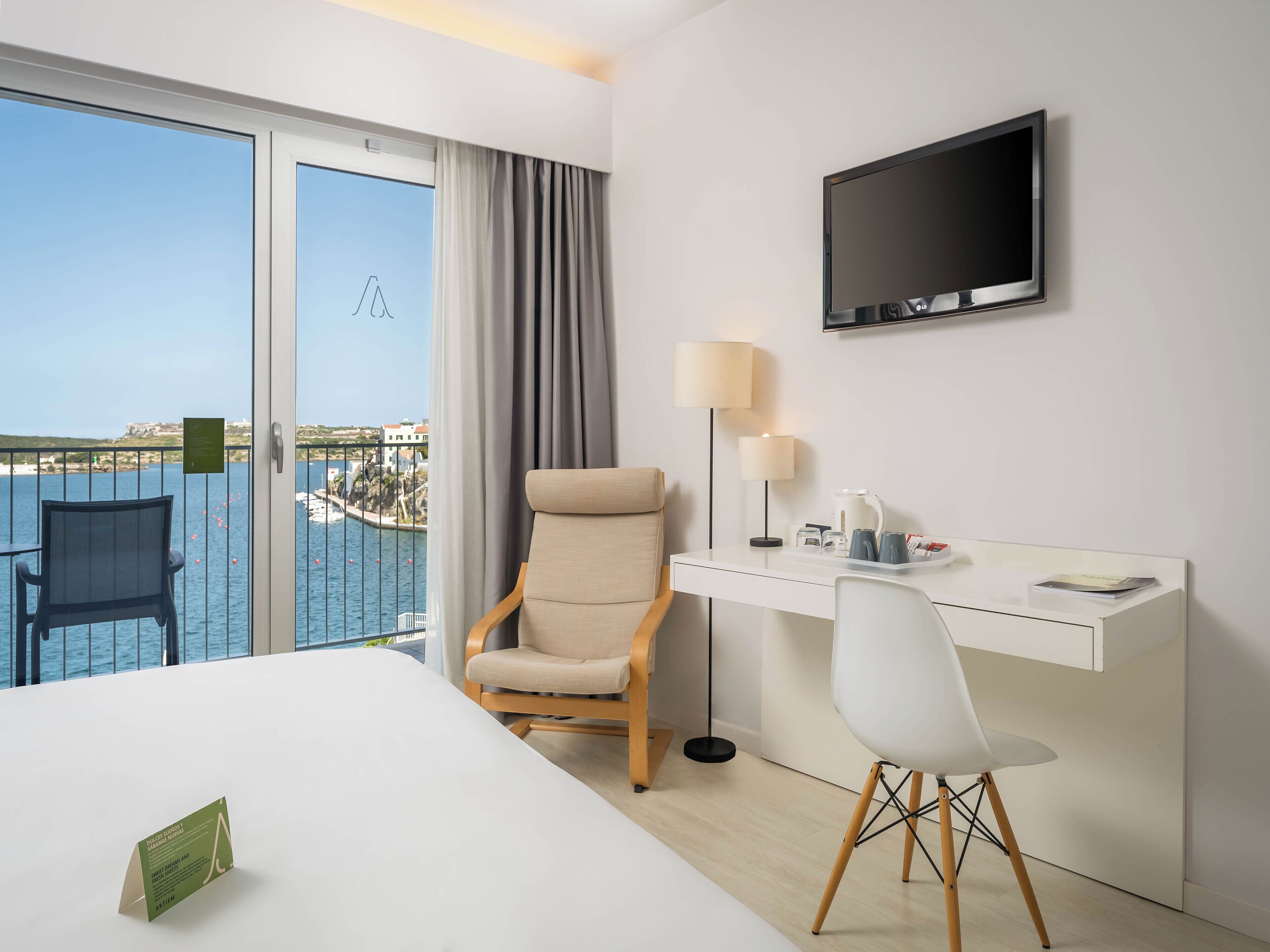 Quarto de hotel com cama, escrivaninha com cadeira, televisão e vista para o mar.