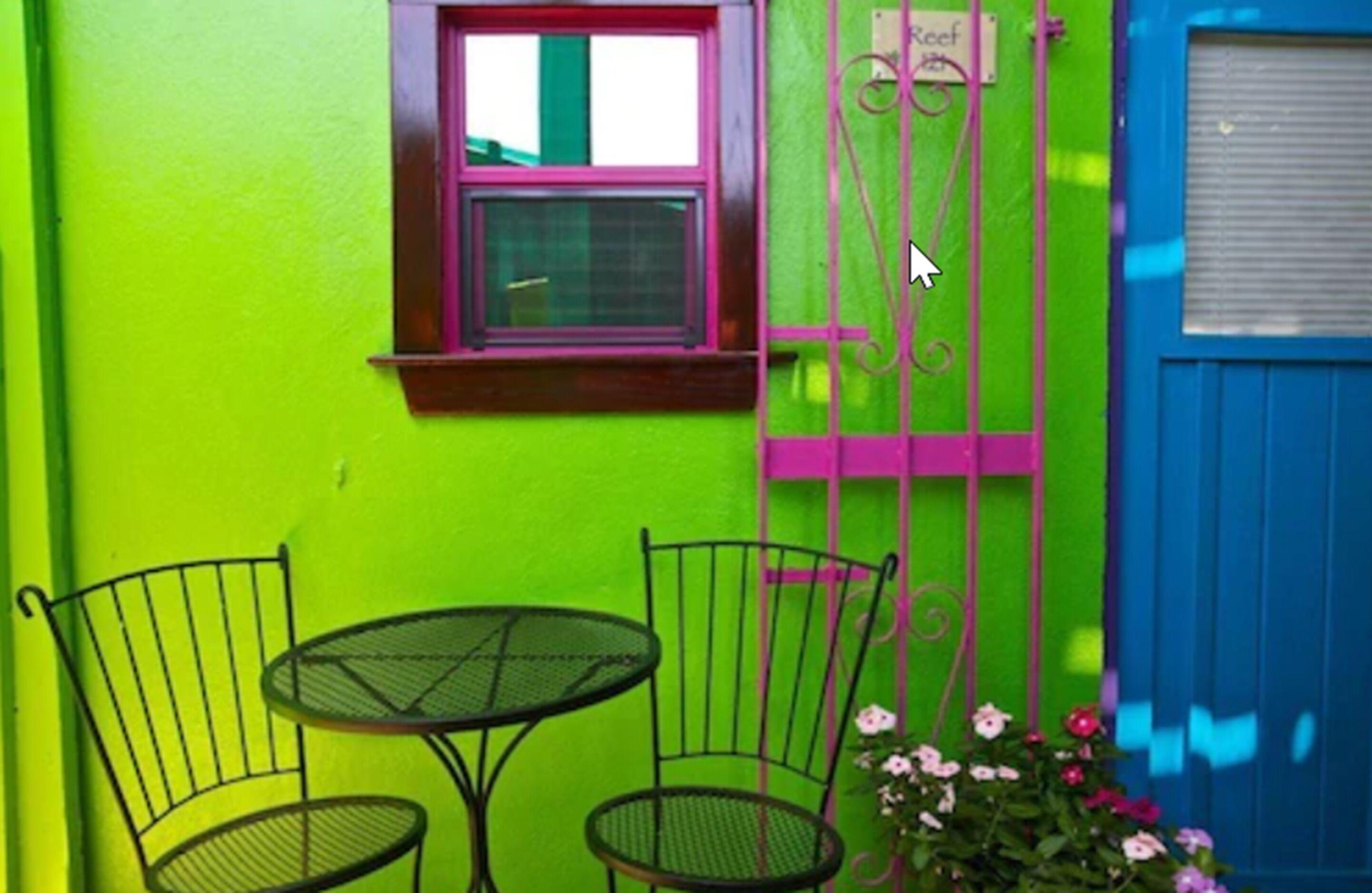 Uma parede verde vibrante com uma janela rosa e uma porta azul. Há duas cadeiras de metal e uma mesa redonda pequena.
