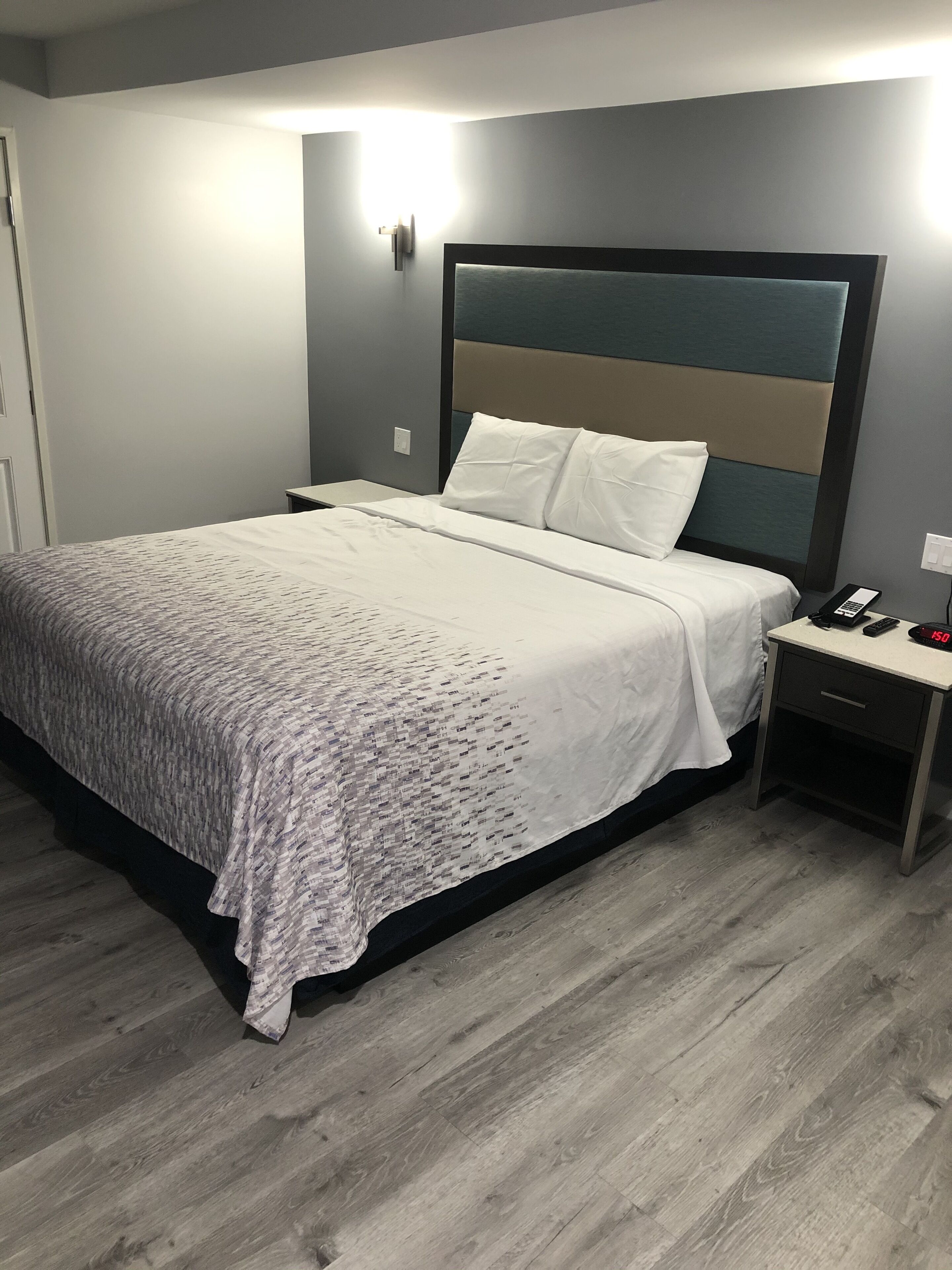 Cama bem arrumada com edredom branco e almofadas, mesa de cabeceira com telefone e relógio, e luminária fixada na parede.