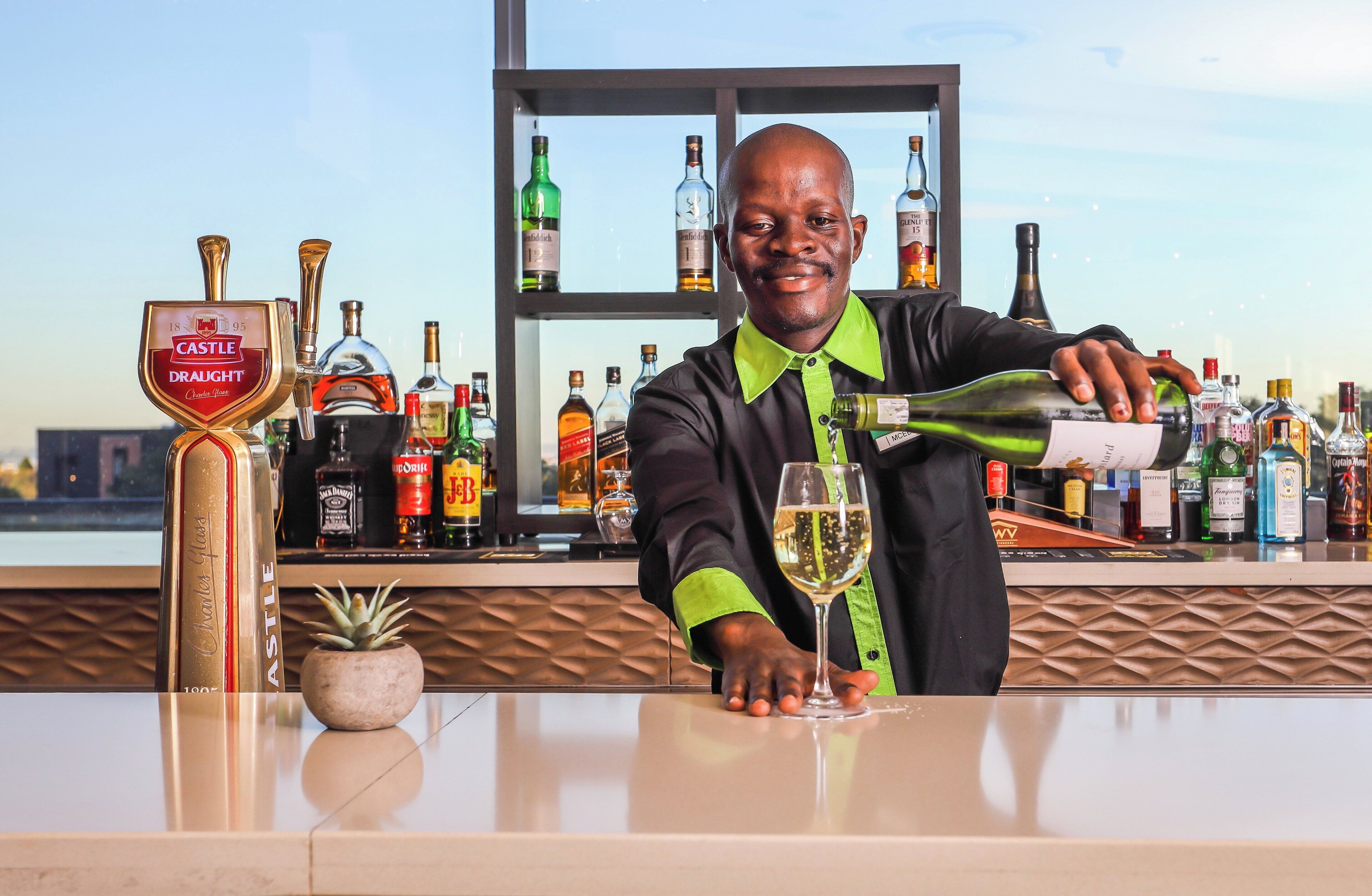Um bartender servindo uma bebida de uma garrafa para um copo em um bar, com diversas garrafas de bebidas alcoólicas ao fundo.