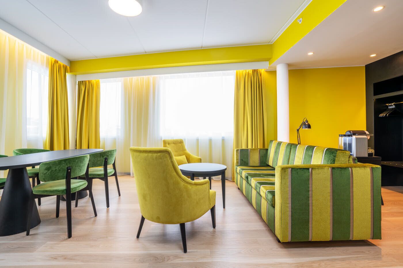 Quarto de hotel moderno com esquema de cores amarelo e verde vibrantes, apresentando uma área de refeições com mesa e cadeiras, um sofá e uma pequena mesa redonda.