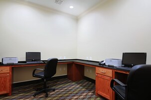 Quarto de hotel com duas mesas, cada uma com um computador e uma impressora, uma cadeira de escritório preta e um tapete estampado.
