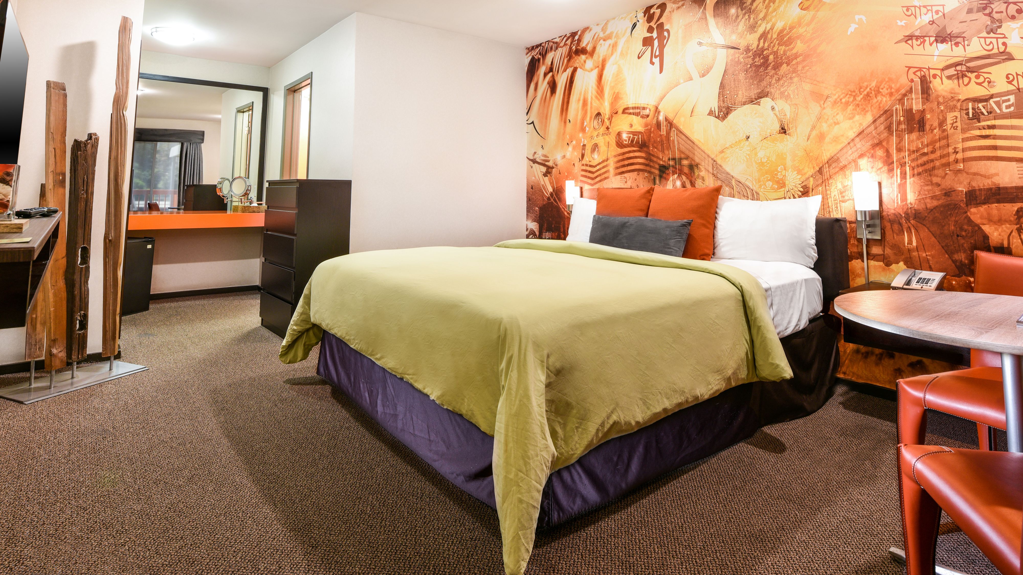Quarto de hotel com cama, escrivaninha com cadeira, espelho e parede com mural.