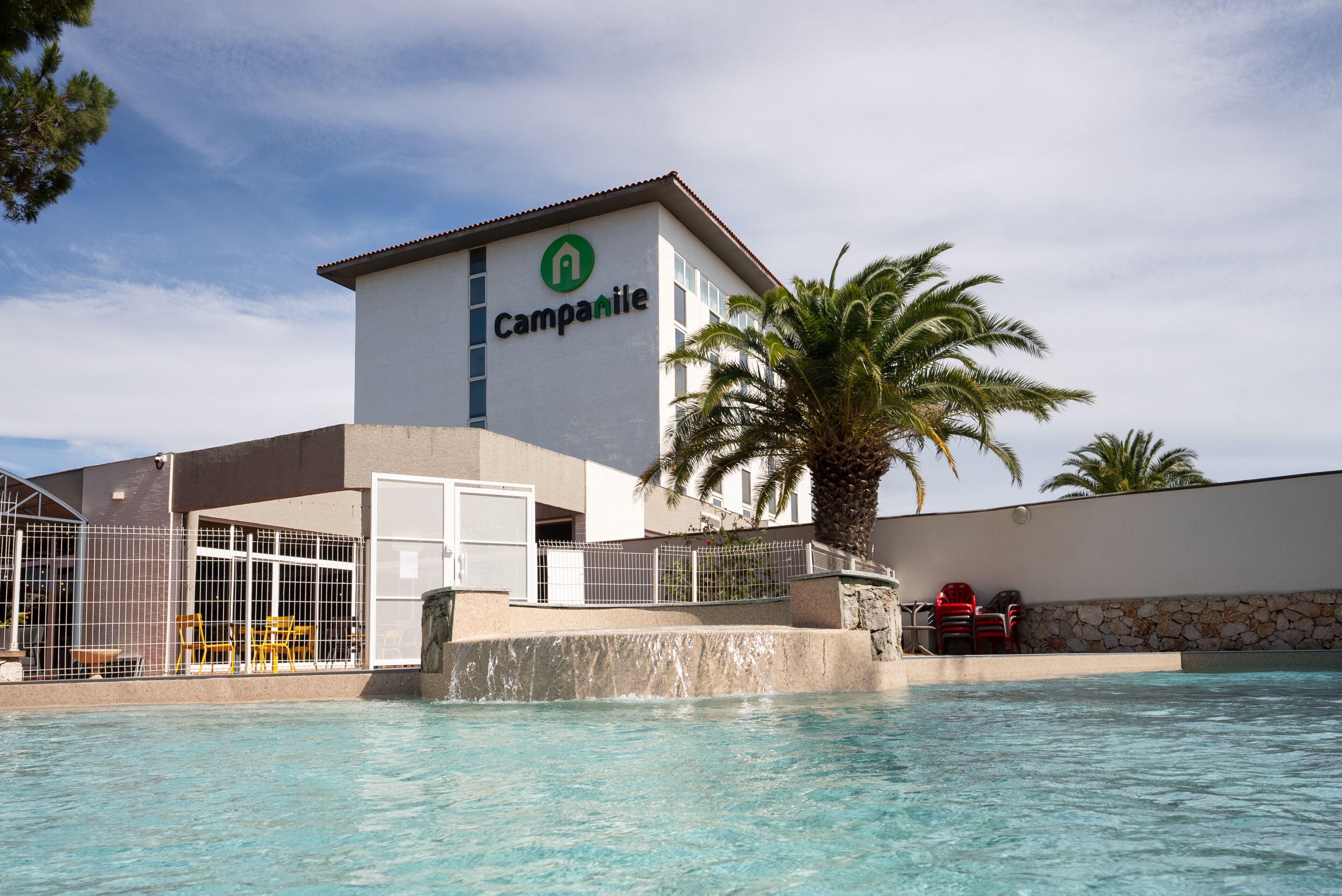 Um edifício com uma placa indicando "Campanile" e uma área de piscina com cadeiras e uma palmeira.