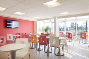 Uma cafeteria bem iluminada, com cadeiras brancas e vermelhas, uma janela ampla e uma televisão de tela plana na parede.