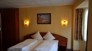 Quarto de hotel com cama de casal, duas mesas de cabeceira, painel de cama de madeira, duas luminárias fixadas na parede e um cartaz com publicidade de férias na Bélgica.