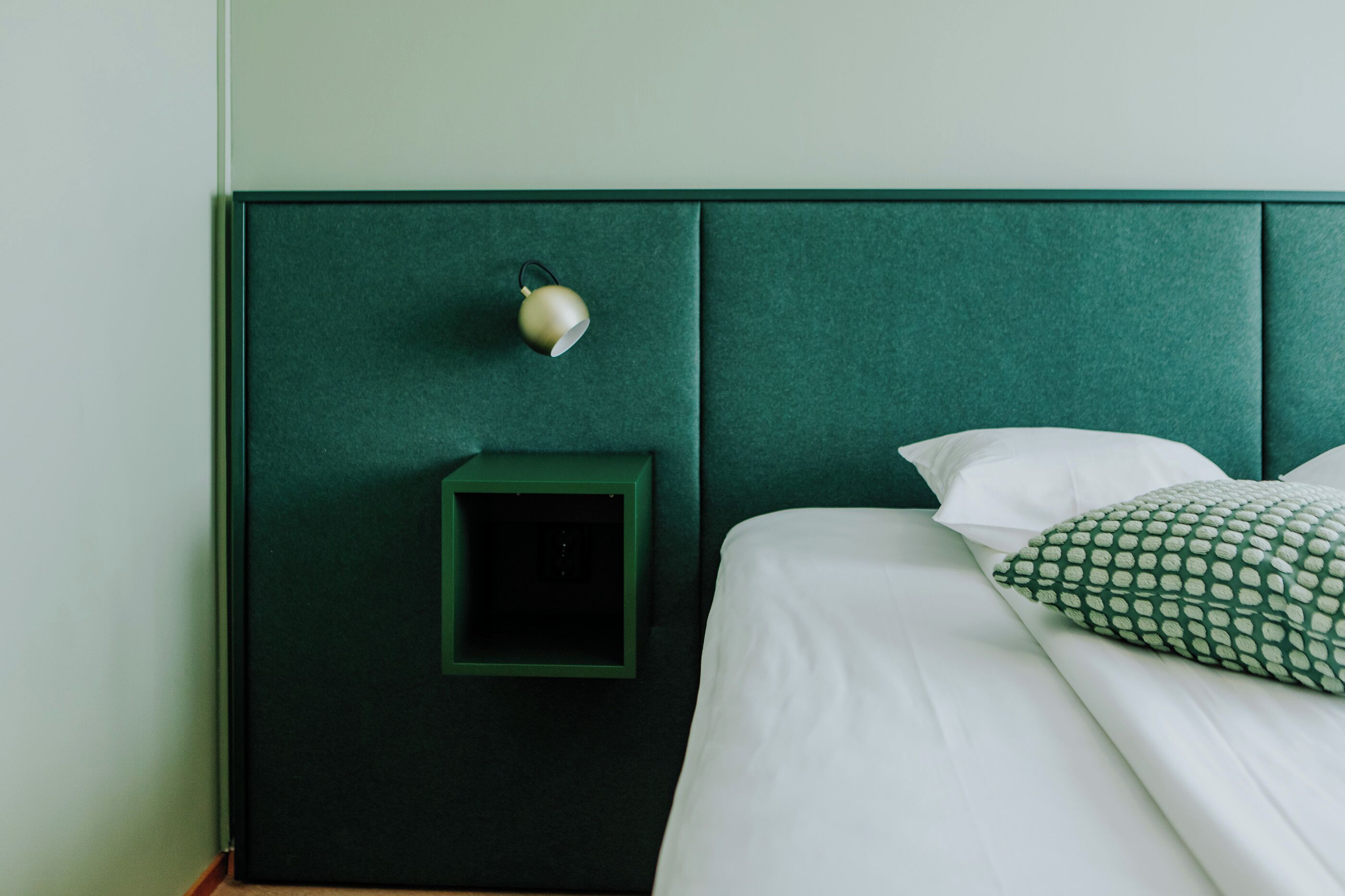 Cama bem arrumada com lençóis brancos e almofada verde estampada, mesa de cabeceira verde com abajur e cabeceira verde.