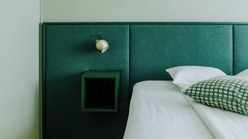 Cama bem arrumada com lençóis brancos e almofada verde estampada, mesa de cabeceira verde com abajur e cabeceira verde.