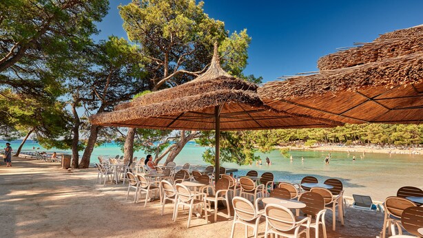 Um café à beira-mar com mesas ao ar livre e guarda-sóis, com vista para um mar azul límpido e uma praia de areia.