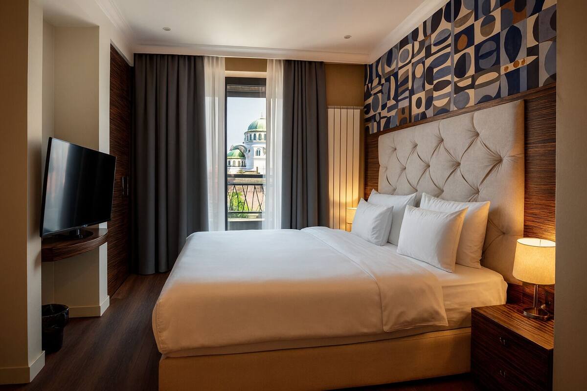 Quarto de hotel com uma cama grande, televisão sobre um suporte de madeira, vista de um edifício pela janela e parede com padrão.