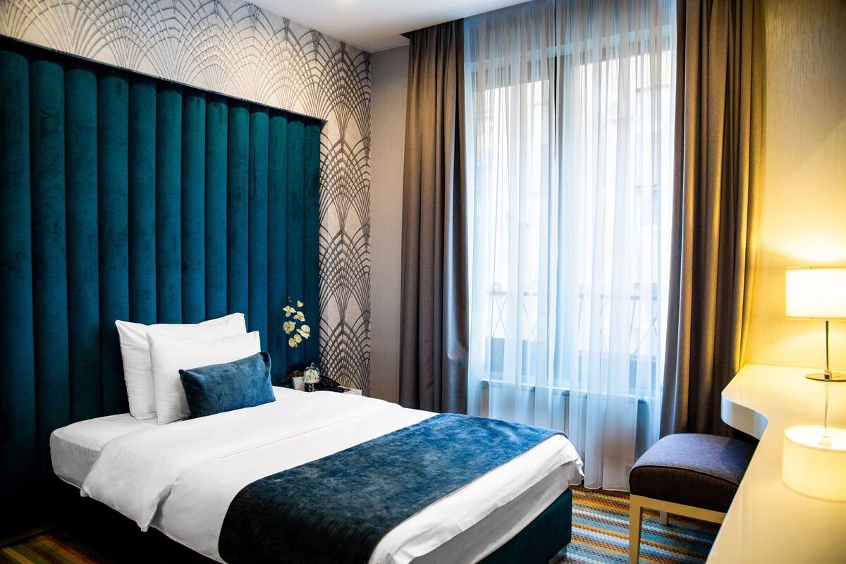 Quarto de hotel com uma cama grande, mesa de cabeceira, cadeira e janela com cortinas.