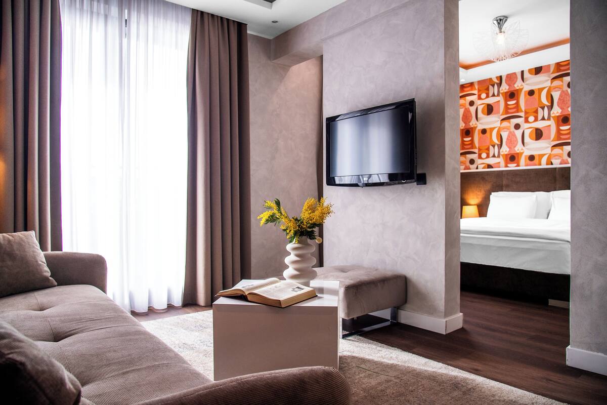Quarto de hotel moderno com sofá, mesa de centro com um livro e flores, TV na parede e cama com cabeceira estampada.