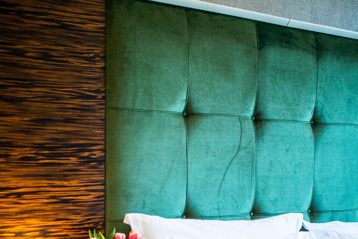 Cama bem arrumada com travesseiros brancos e rosa, cabeceira estofada em verde, mesa de cabeceira com abajur e um vaso com flores.