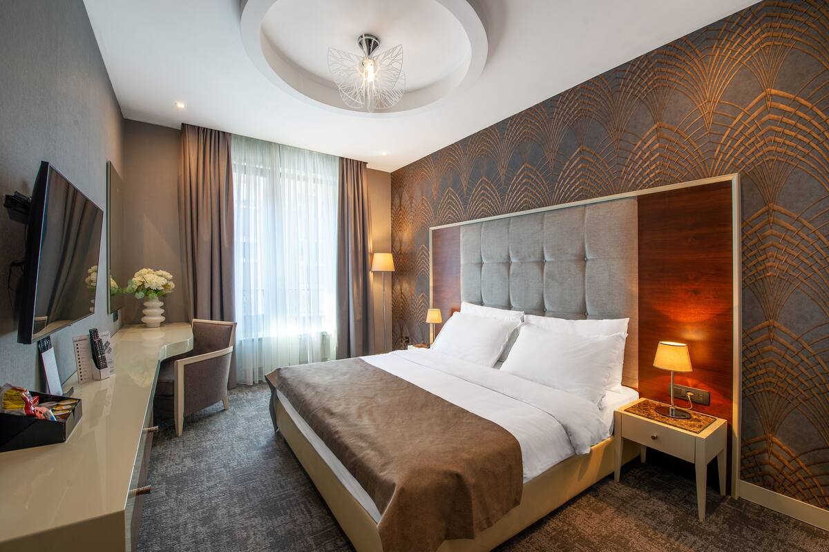 Quarto de hotel com uma cama grande, uma mesa com televisão de tela plana, uma mesa de cabeceira com abajur e uma parede decorativa.