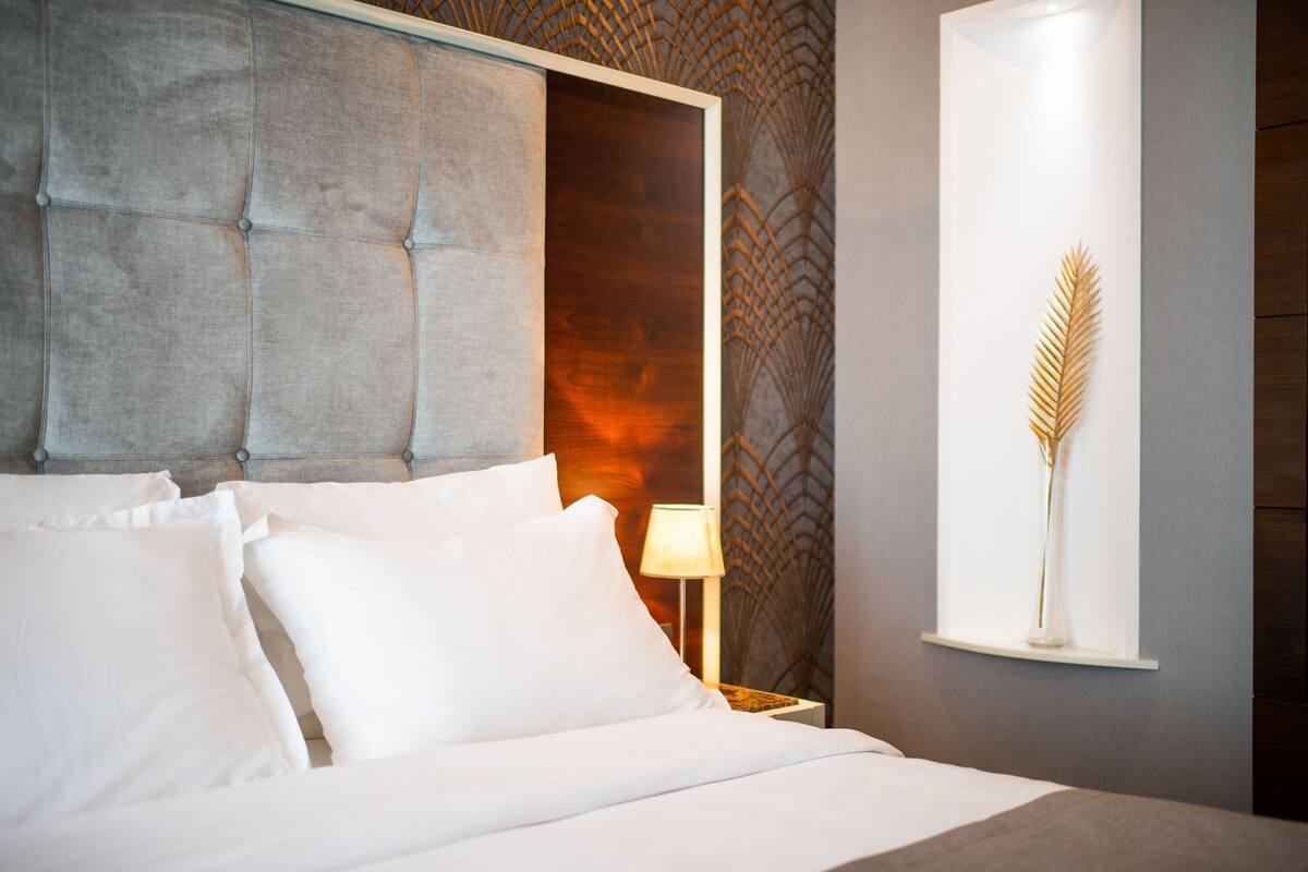 Uma cama bem arrumada com lençóis e almofadas brancas, uma cabeceira estofada, um abajur e um objeto decorativo na parede.