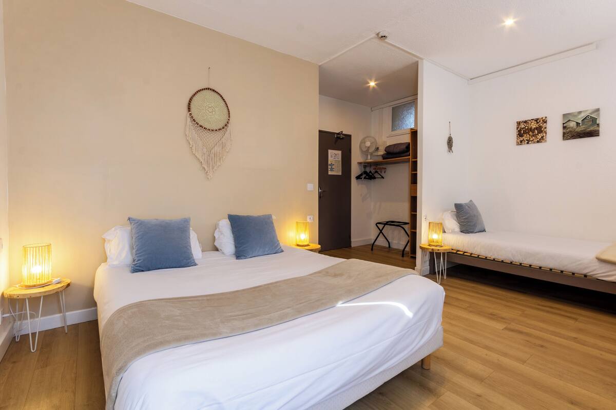 Um quarto com uma cama grande, uma cama menor, uma mesa de cabeceira com abajur, um enfeite na parede e uma porta que dá para outro cômodo.