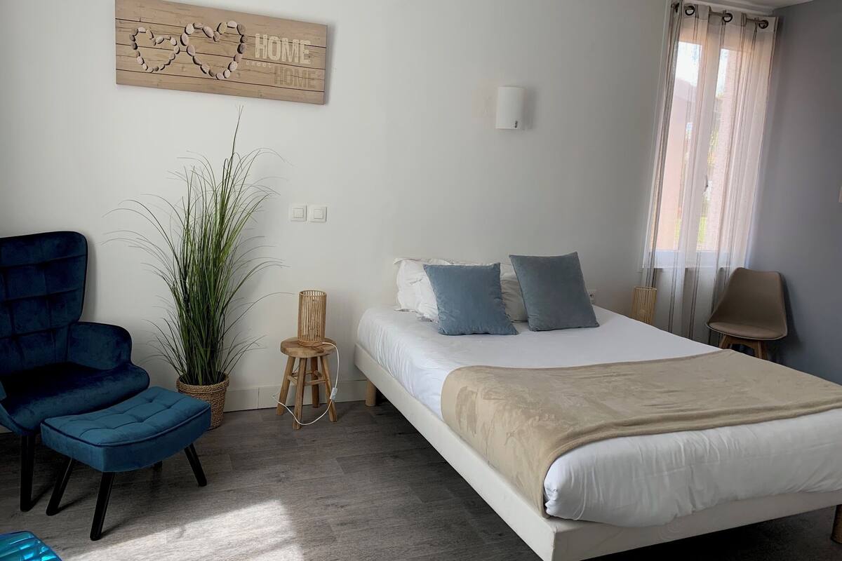 Quarto com cama, uma cadeira azul, planta em vaso e luminária na parede.