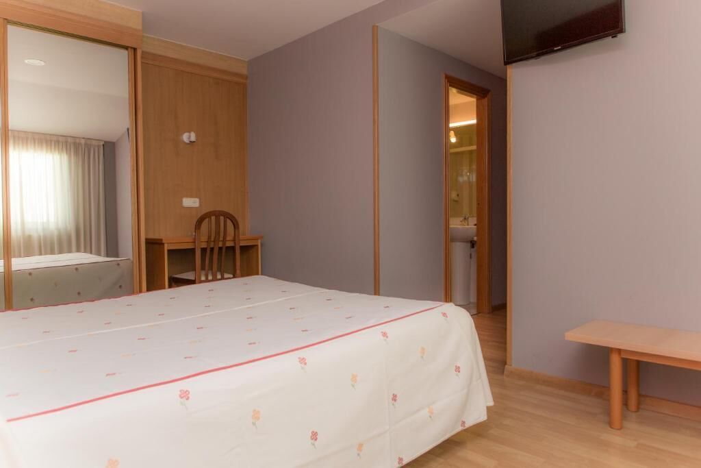 Quarto com cama, escrivaninha de madeira, cadeira, televisão e banheiro visível através de uma porta aberta.
