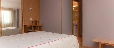 Quarto com cama, escrivaninha de madeira, cadeira, televisão e banheiro visível através de uma porta aberta.