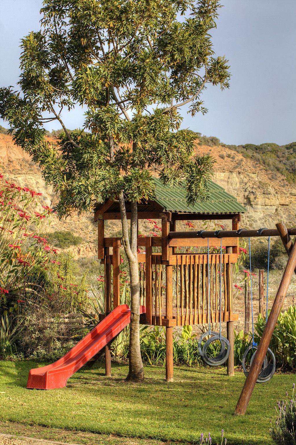 Uma casinha de brincar de madeira com um escorrega vermelho, rodeada por vegetação e uma paisagem ondulada.