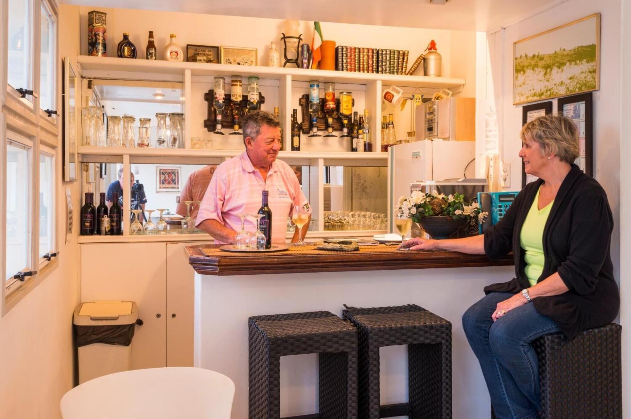 Uma área de bar com prateleiras de garrafas e copos, um balcão e banquetas de vime.