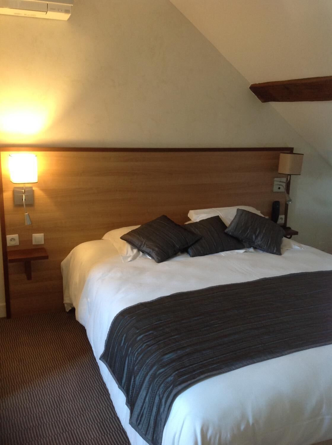 Quarto de hotel com uma cama grande, cabeceira de madeira, mesa de cabeceira, abajur e luminária na parede.