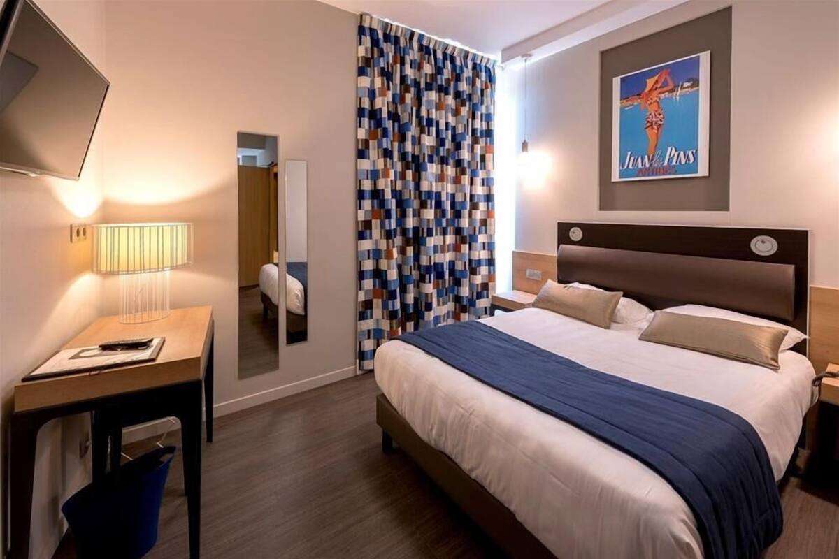 Quarto de hotel com cama, mesa de cabeceira, abajur, espelho e um quadro na parede.