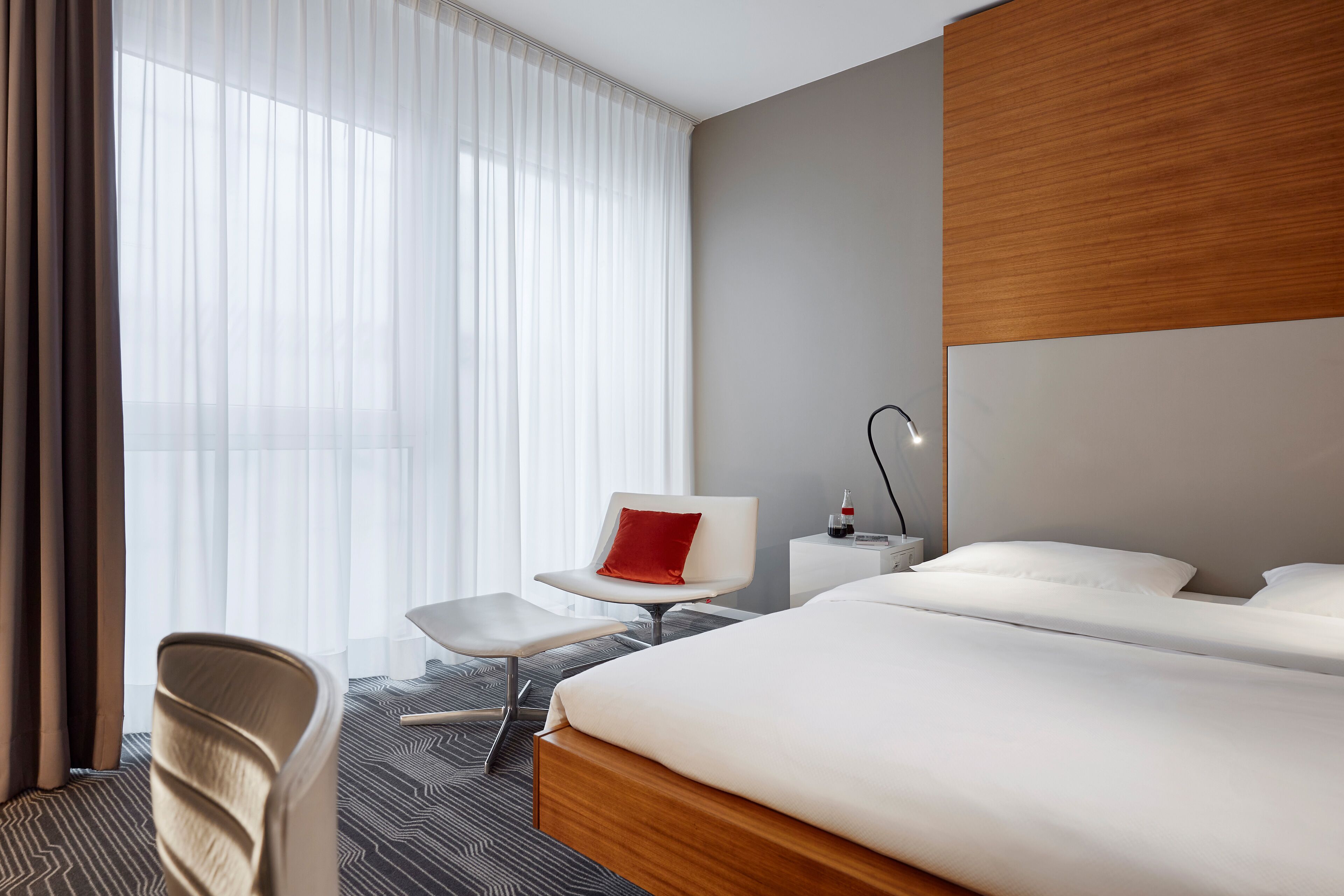 Quarto de hotel moderno com uma cama grande, uma escrivaninha, uma cadeira e uma janela com cortinas leves.