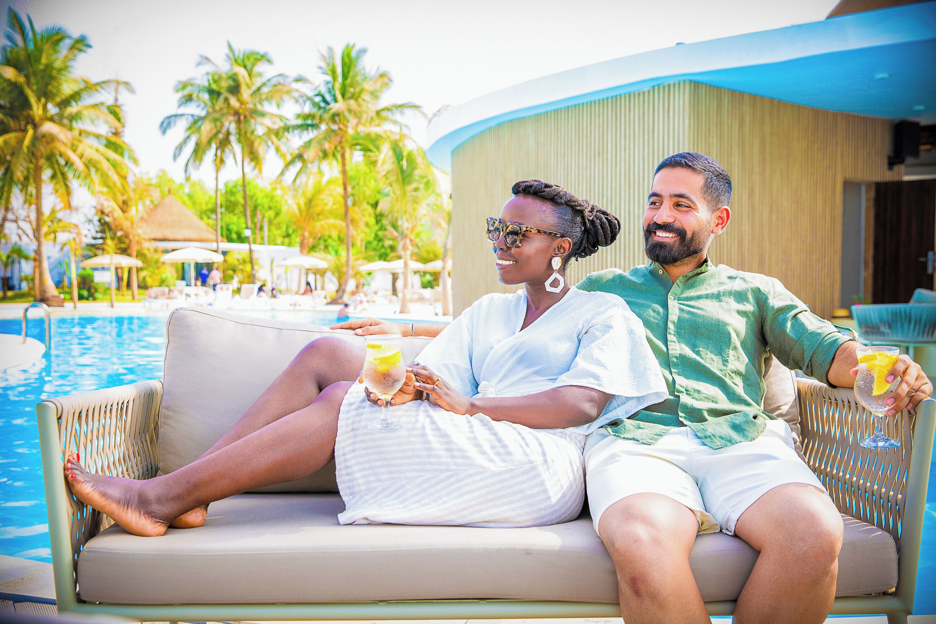 Um casal relaxando em uma espreguiçadeira à beira da piscina, com vista para palmeiras e um edifício ao fundo.