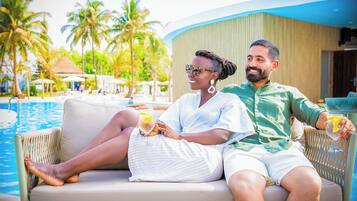 Um casal relaxando em uma espreguiçadeira à beira da piscina, com vista para palmeiras e um edifício ao fundo.