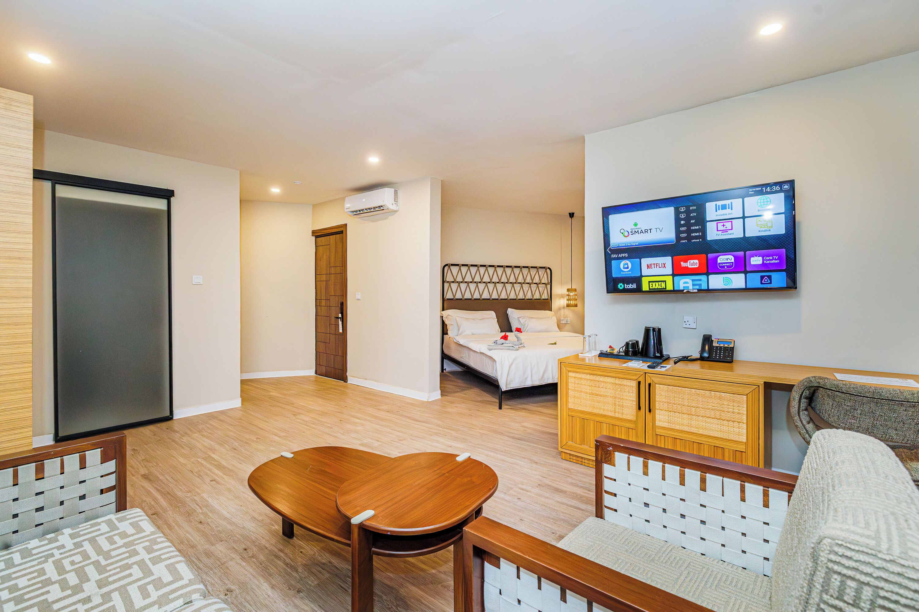 Quarto de hotel moderno com cama, televisão fixada na parede exibindo vários canais, escrivaninha de madeira e área de estar com poltrona e sofá.