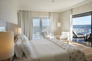 Quarto de hotel com uma cama grande, área de estar com uma cadeira e mesa, e varanda com vista para a marina.
