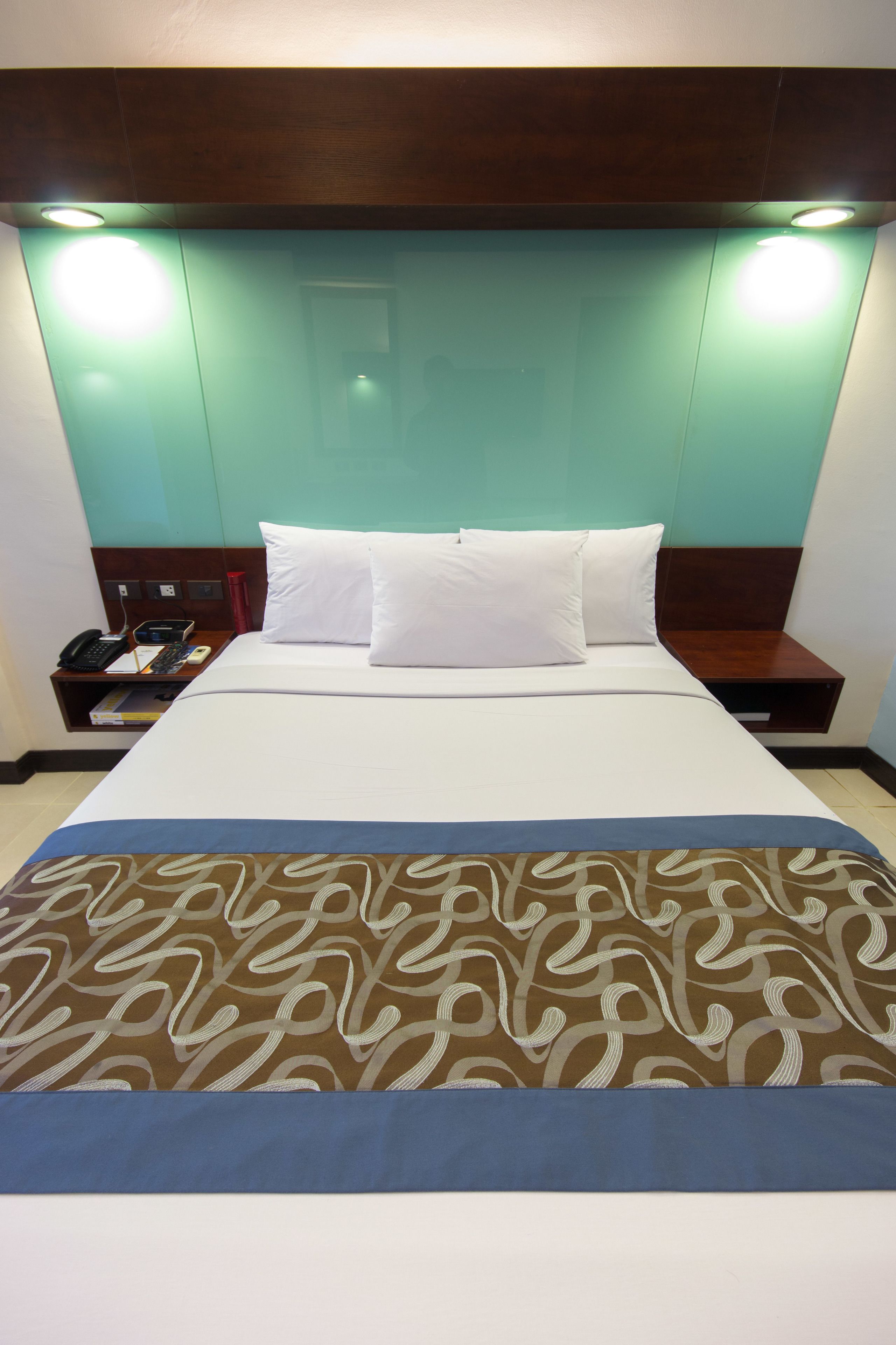 Quarto de hotel com cama bem arrumada, mesa de cabeceira com telefone e um livro, e cabeceira com iluminação embutida.