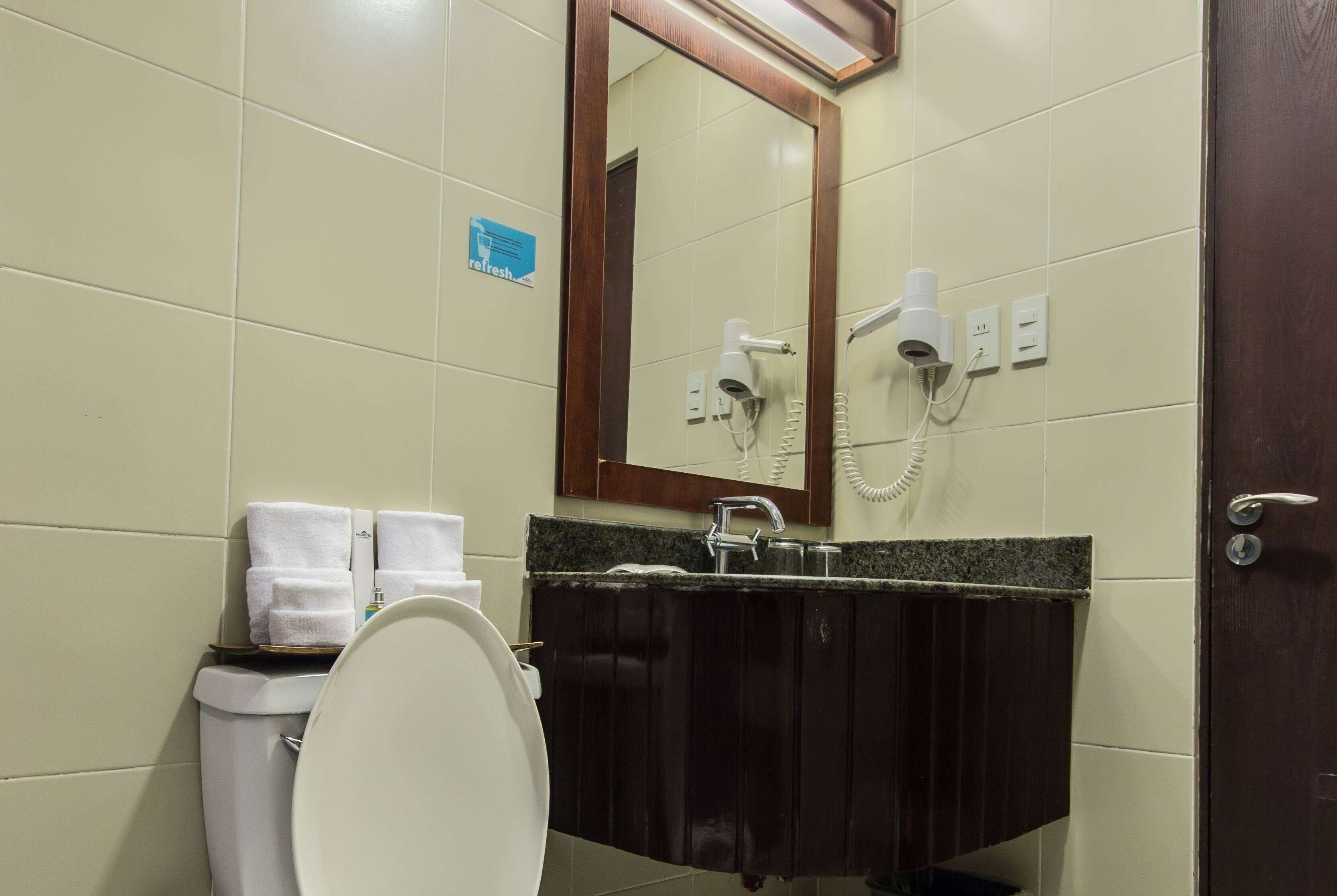 Um banheiro com vaso sanitário, pia com espelho acima e secador de cabelo fixado na parede.