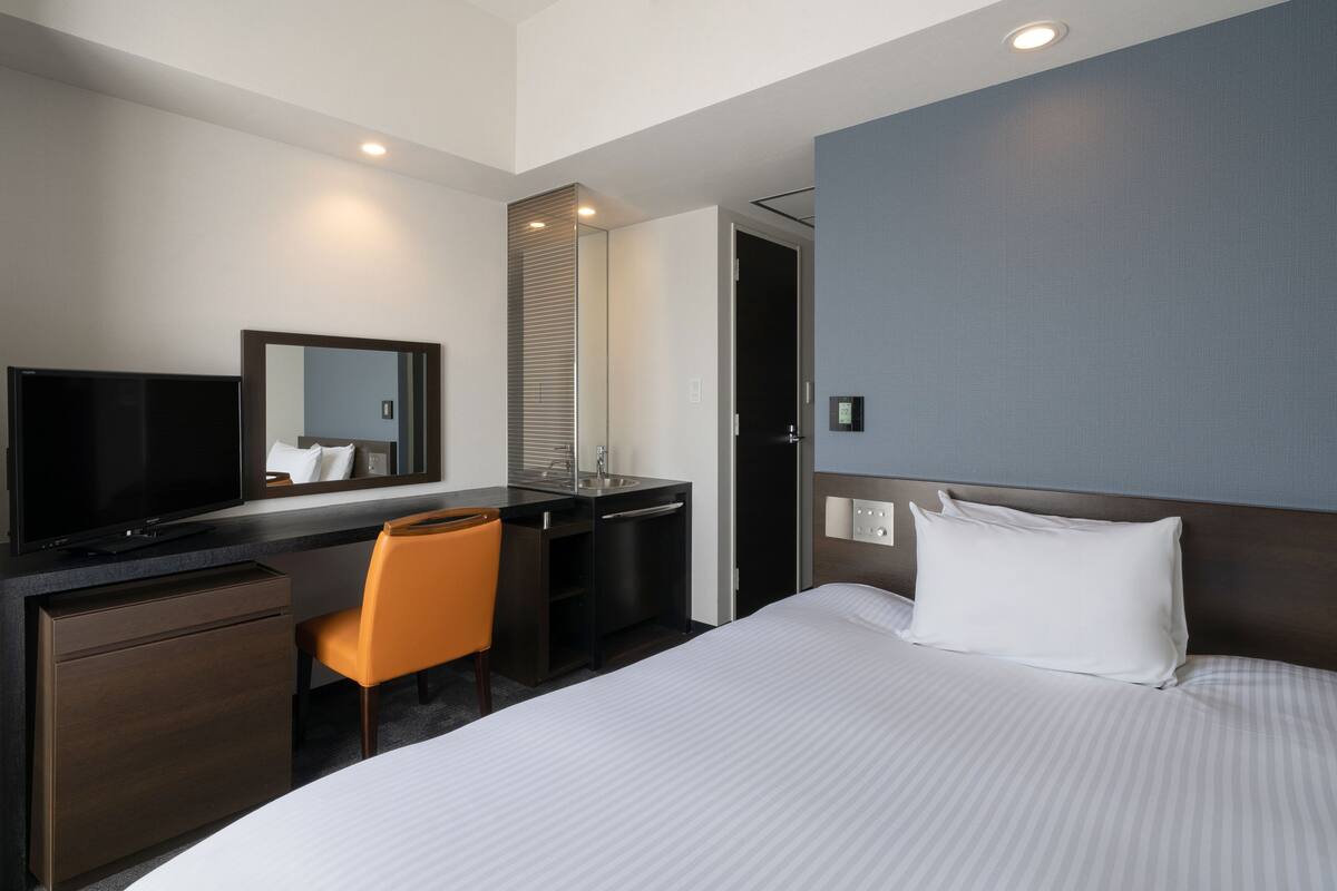 Quarto de hotel com uma cama, uma escrivaninha com cadeira, televisão, espelho e porta do banheiro.