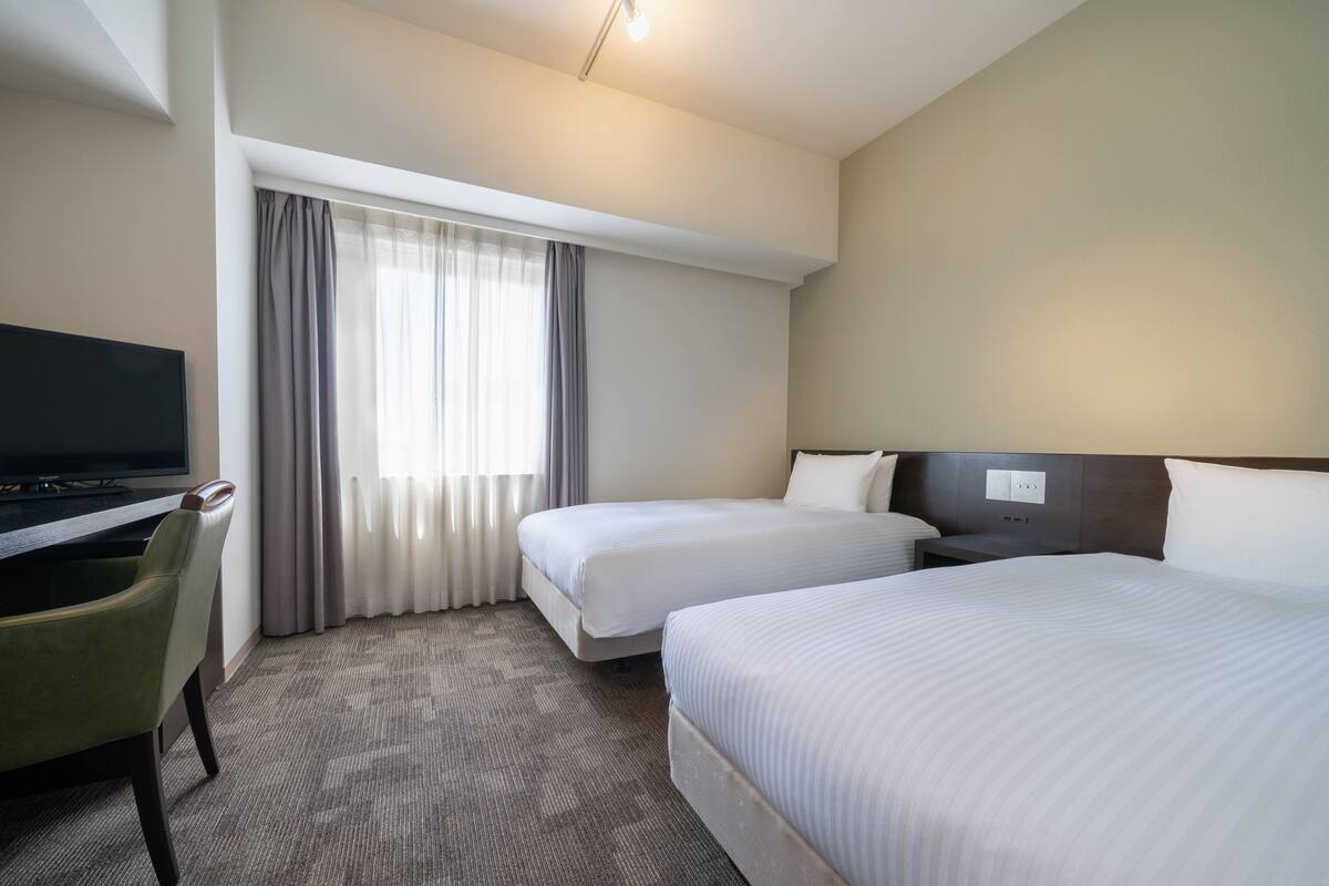 Quarto de hotel com duas camas, uma escrivaninha, televisão e janela com cortinas.