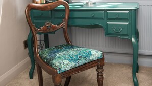 Uma cômoda azul-turquesa com espelho, uma cadeira de madeira com almofada estampada e papel de parede floral.