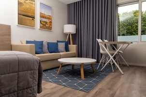 Quarto de hotel moderno com sofá, mesa redonda, luminária e vista para o exterior.