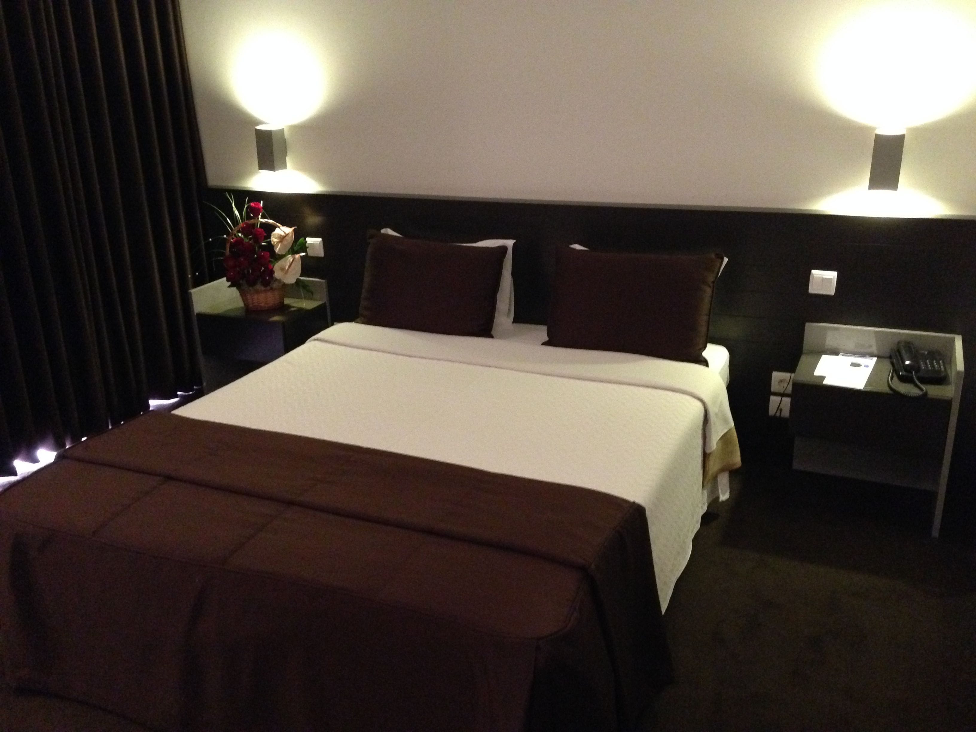 Quarto de hotel com uma cama grande, duas mesas de cabeceira, um criado-mudo, um abajur e um telefone.