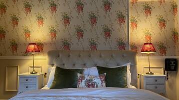 Quarto com cabeceira estofada, duas mesas de cabeceira com abajures e papel de parede floral.