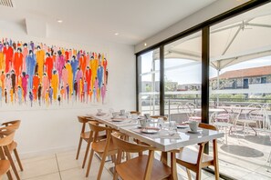 Uma sala de jantar com uma mesa comprida posta para seis pessoas, rodeada por cadeiras de madeira, e um quadro abstrato e colorido na parede.
