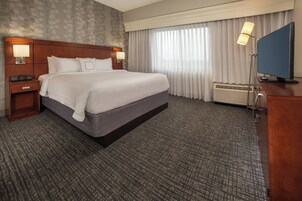 Quarto de hotel com uma cama grande, uma escrivaninha com televisão de tela plana, uma mesa de cabeceira com luminária e uma janela com cortinas.