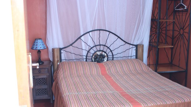 Uma cama com colcha listrada e dossel, uma mesa de cabeceira com abajur e uma estante de metal contra uma parede cor de pêssego.