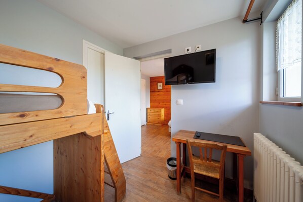 Quarto com cama de madeira, televisão na parede, uma mesinha com cadeira e radiador.