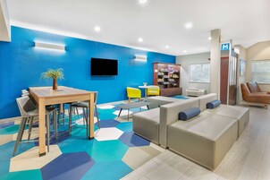 Quarto de hotel moderno com uma parede de destaque azul, televisão de tela plana, área de refeições com mesa e cadeiras, um sofá e uma estante de livros.