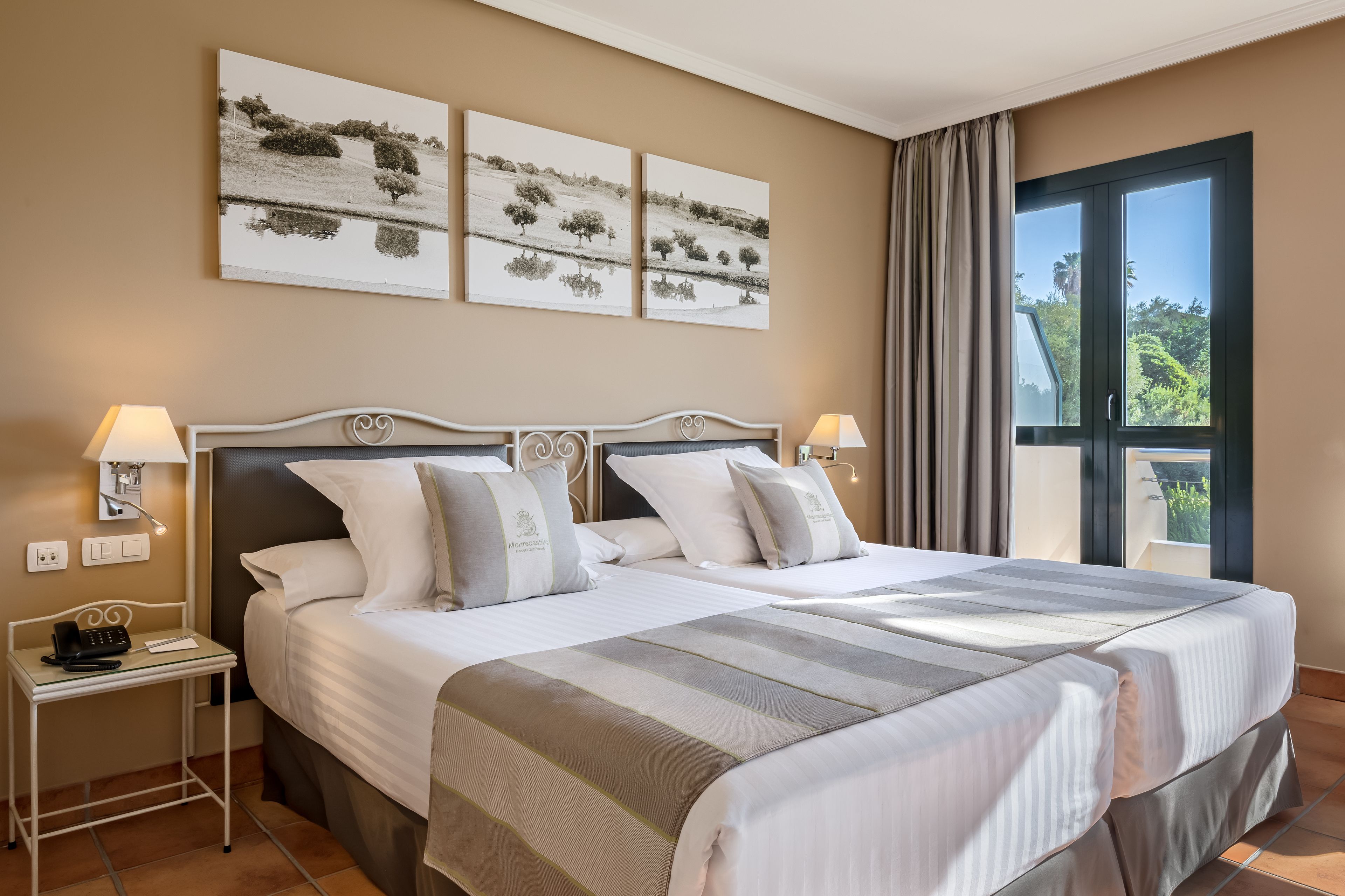 Uma cama de casal com colcha listrada, duas luminárias de cabeceira, mesa de cabeceira com telefone e obra de arte na parede retratando paisagens.