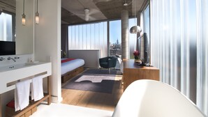 Quarto de hotel moderno com uma cama grande, uma mesa de trabalho, uma cadeira, uma televisão e uma janela ampla com vista para a cidade.