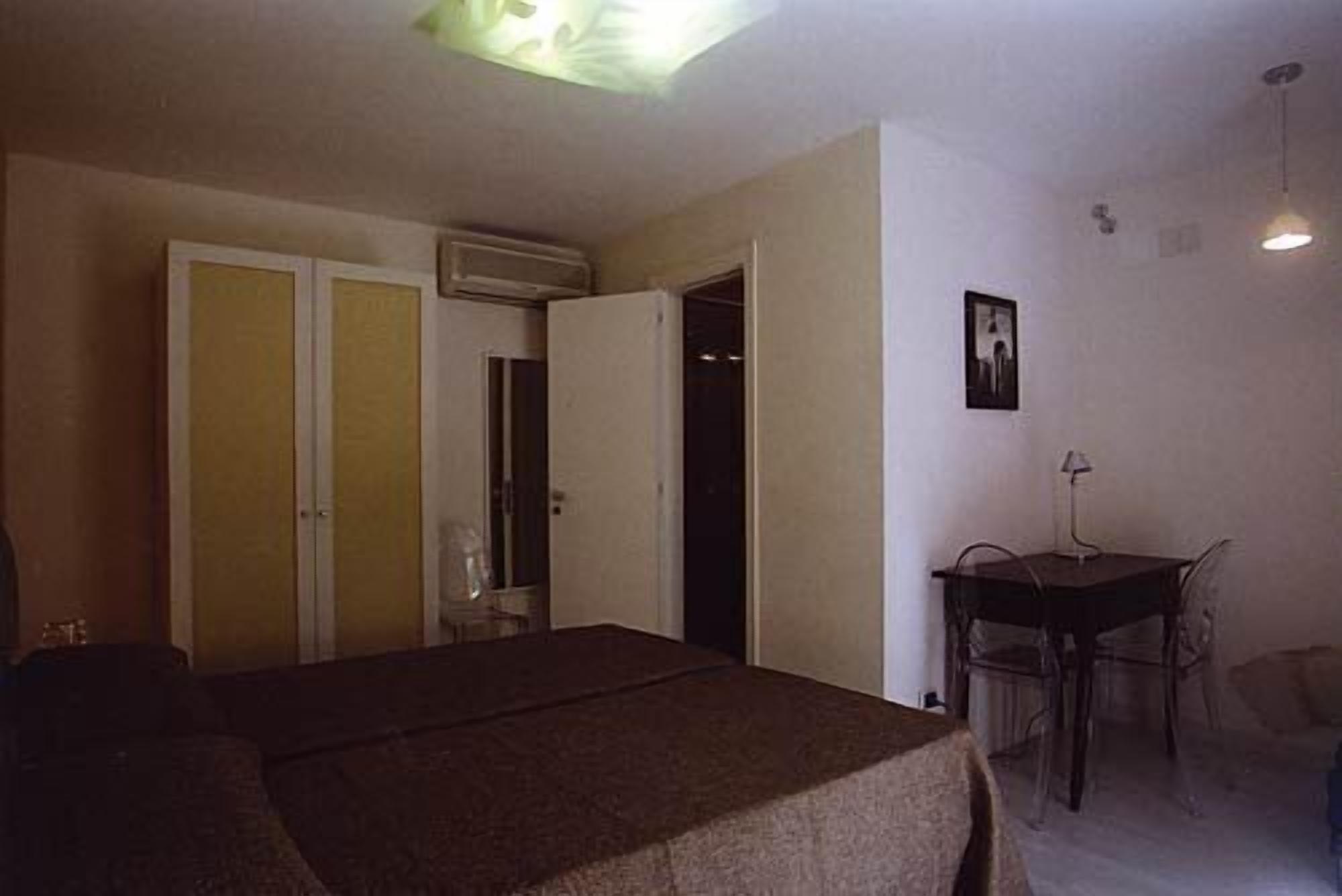Quarto com cama, escrivaninha e cadeira. Há uma porta que dá para outro cômodo.