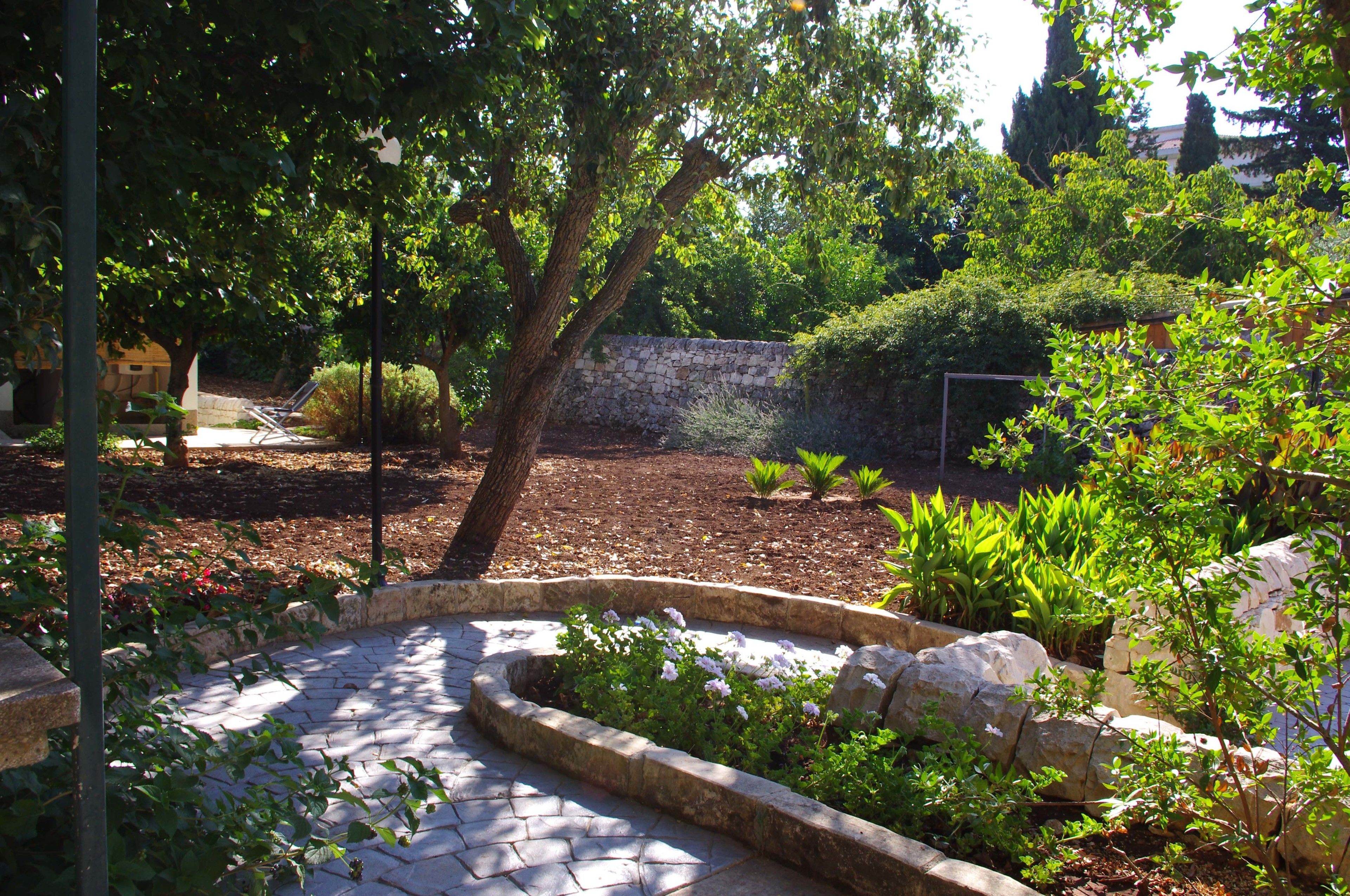 Área ajardinada pavimentada, com muro de pedra, diversas plantas e um banco.